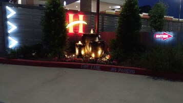 Entrada de hotel com placa de 'Fire Lane', placa de 'Não Estacionar' e um grande letreiro iluminado em formato de 'H'.