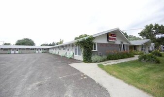Um motel com uma fileira de edifícios térreos, estacionamento e uma área gramada com calçada.