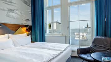 Quarto de hotel com cama, poltrona, mesa, luminária e vista da cidade por grandes janelas.