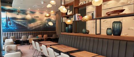 Um restaurante com mesas e cadeiras de madeira, iluminação pendente e um mural de um veleiro na parede.