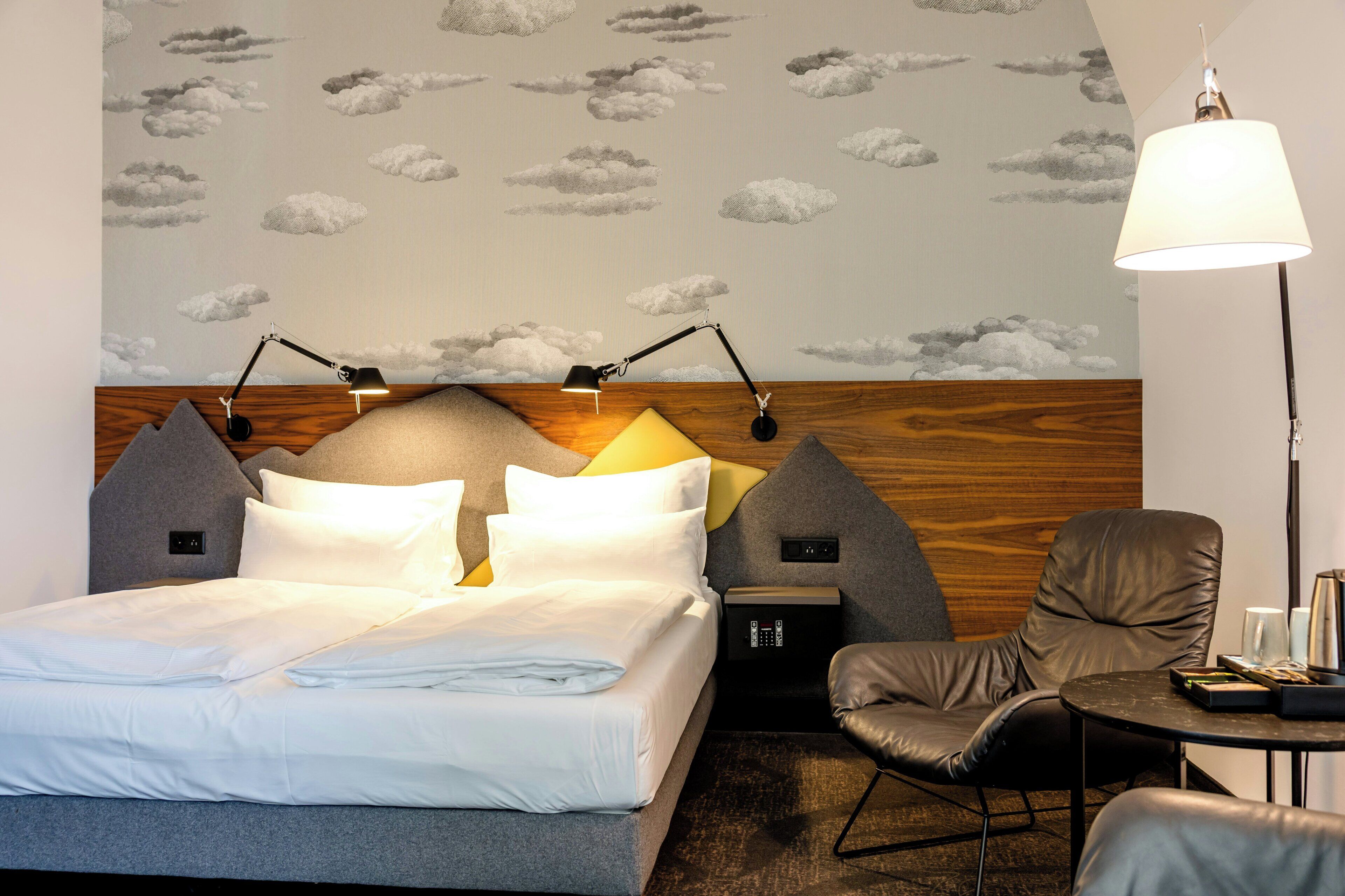 Quarto de hotel com cama, luminárias laterais, uma cadeira, uma mesa e parede com padrão de nuvens.