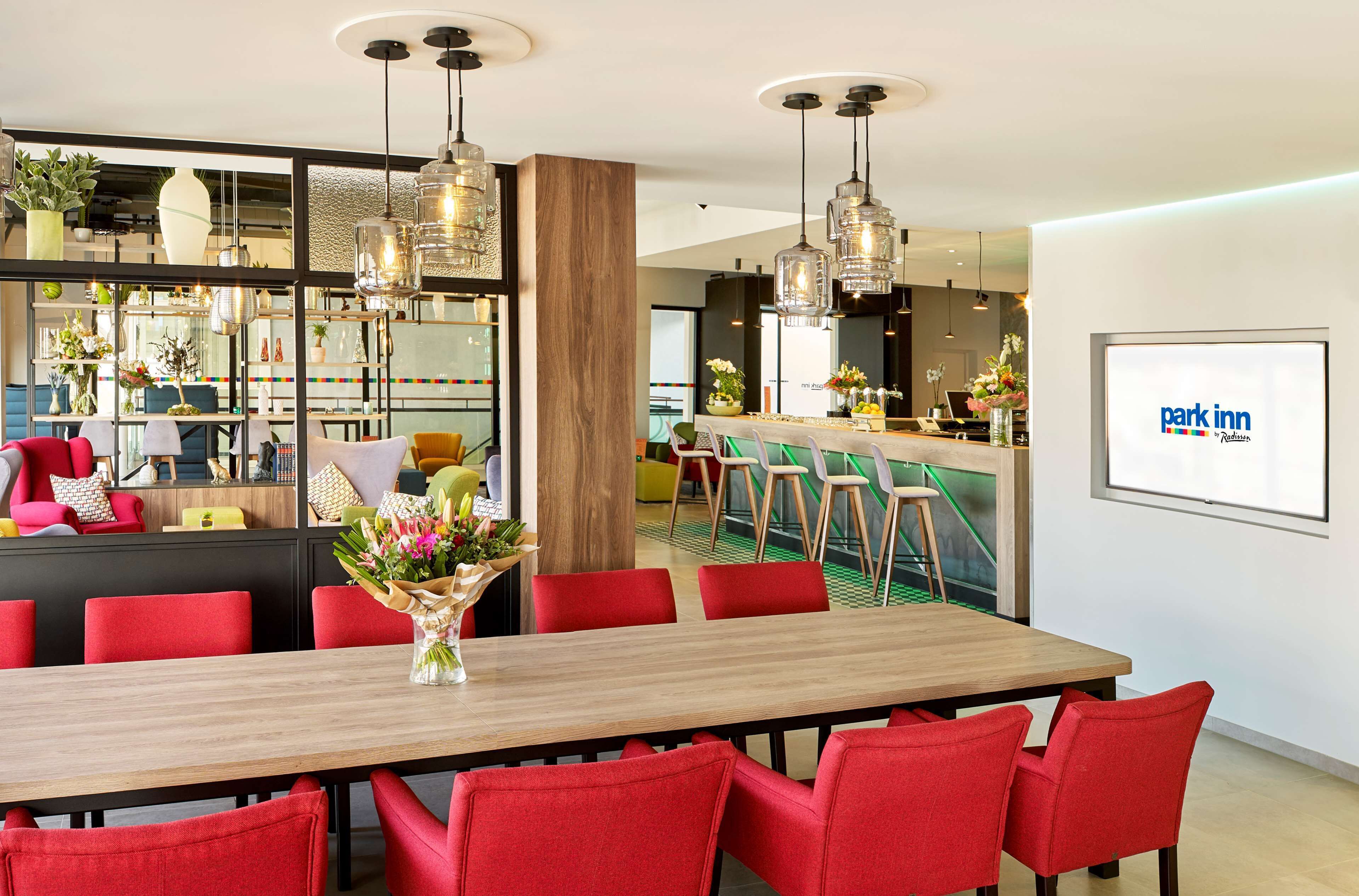 Uma sala de jantar de hotel moderna, com uma longa mesa de madeira, cadeiras vermelhas e luminárias pendentes.