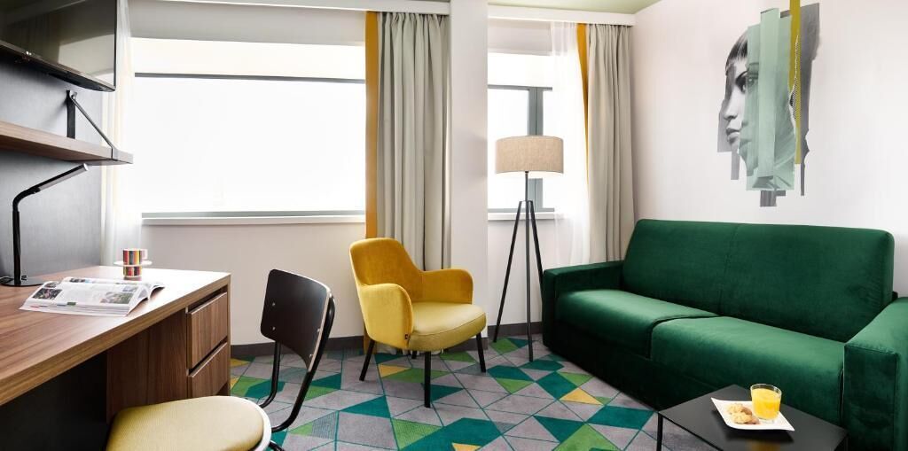 Quarto de hotel com escrivaninha, duas cadeiras, um sofá verde, luminária de chão e uma obra de arte na parede.