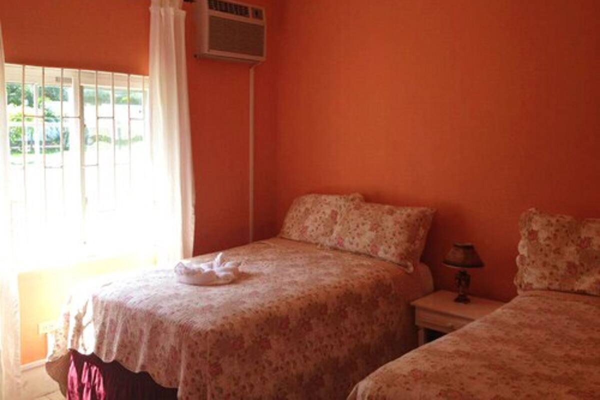 Quarto com duas camas, janela com grades, ar condicionado e abajur sobre uma mesa de cabeceira.
