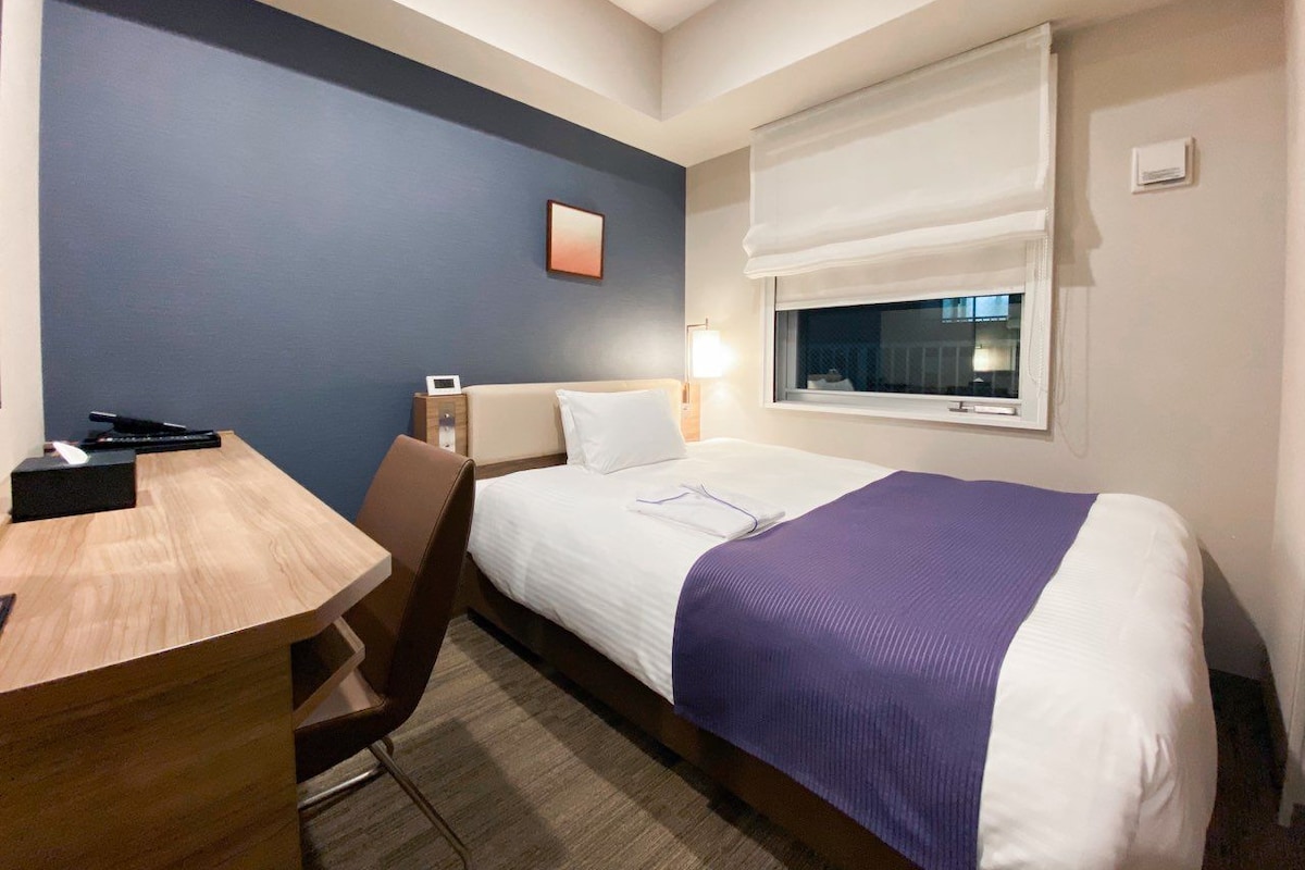 Quarto de hotel com cama, escrivaninha, cadeira, mesa de cabeceira e janela com cortina branca.