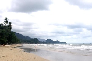 Uma praia com costa arenosa, palmeiras e paisagem montanhosa ao fundo.