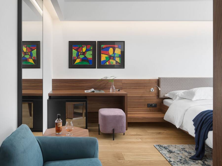 Quarto de hotel moderno com escrivaninha de madeira, sofá confortável, cama com roupa de cama branca e arte abstrata na parede.