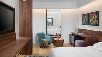 Quarto de hotel moderno com uma cama grande, área de estar com uma poltrona azul, uma pequena escrivaninha e uma televisão.