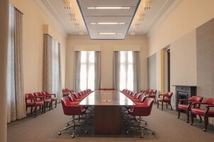 Sala de conferência com uma mesa comprida e cadeiras vermelhas, grandes janelas com cortinas e uma lareira.