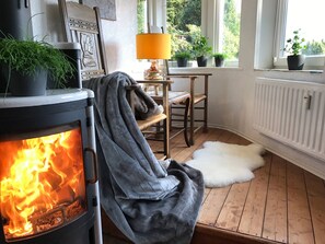 Um quarto acolhedor com lareira a lenha, uma mesa com abajur e algumas plantas em vasos junto à janela.