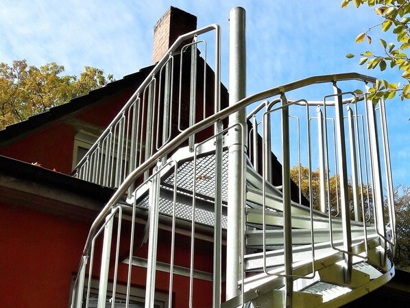 Uma escadaria espiral de metal com corrimão, que leva a um edifício com fachada vermelha e chaminé.