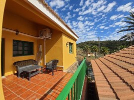 Um edifício amarelo com um pátio pavimentado, guarda-corpo verde e vista para uma paisagem montanhosa.