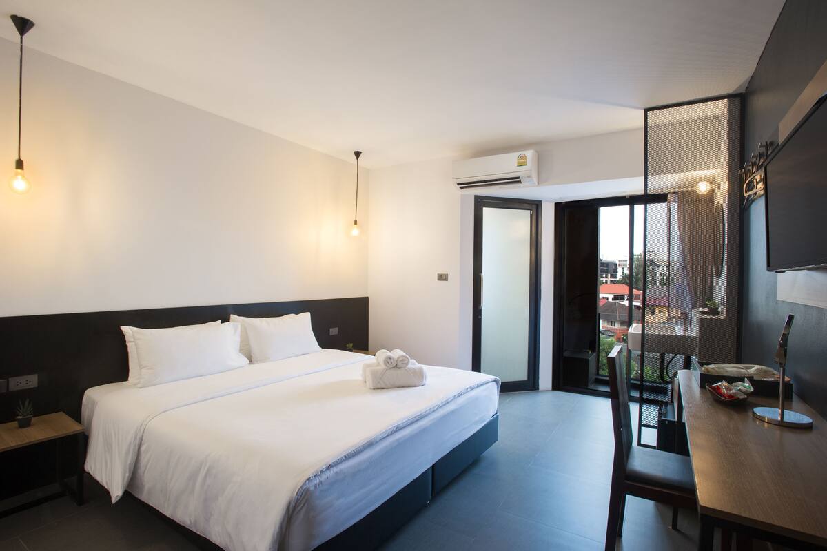 Quarto de hotel moderno com uma cama grande, uma escrivaninha com cadeira e vista para a cidade através de uma porta de varanda.