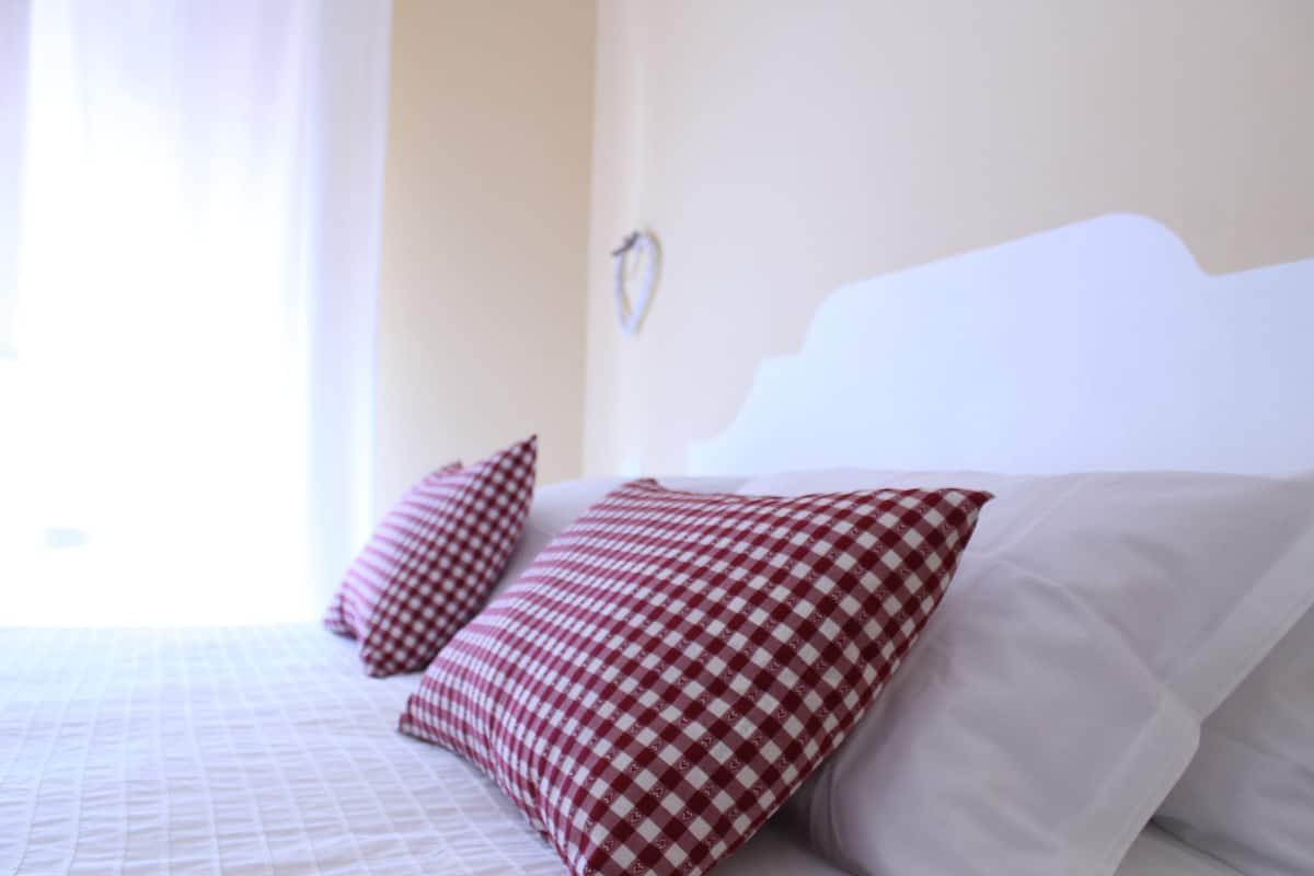 Cama bem arrumada com travesseiro xadrez e um travesseiro liso, janela com cortinas leves e luminária fixada na parede.