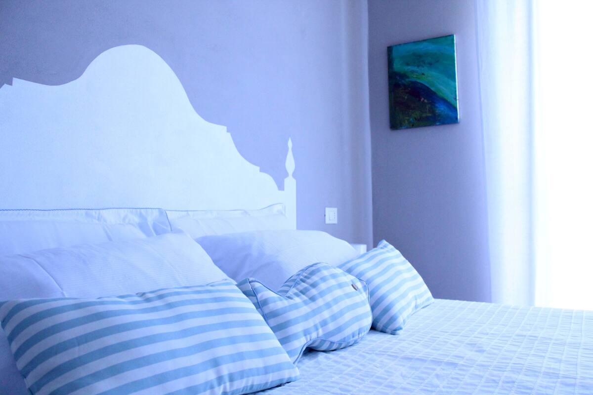 Uma cama com almofadas listradas e cabeceira, um quadro na parede e uma janela com luz natural.