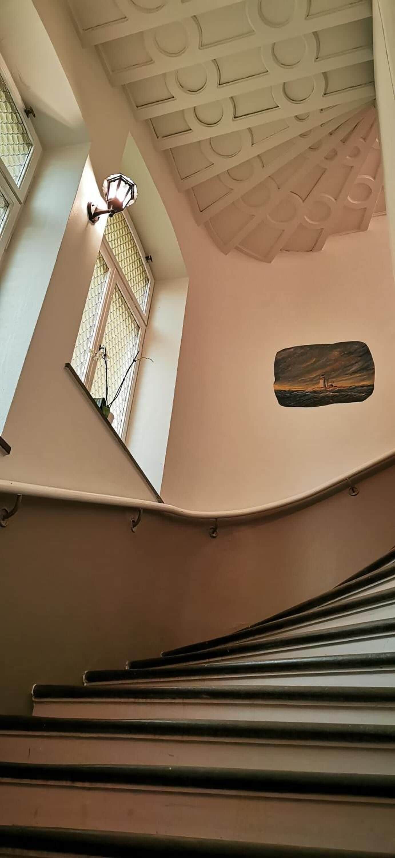 Uma escadaria com corrimão curvo, teto decorativo e uma pequena pintura de um pôr do sol na parede.