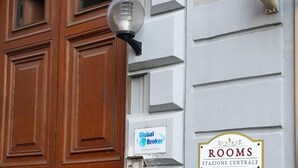 Entrada de um edifício com porta de madeira, câmera de segurança, luminária e placa com a inscrição "Global Broker Rooms" na parede.