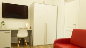 Quarto com guarda-roupa branco, escrivaninha com cadeira, sofá vermelho e televisão de tela plana fixada na parede.