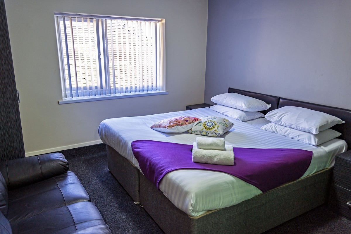 Cama bem arrumada com manta roxa, almofadas brancas e uma almofada floral. Há uma janela com persianas, um banco de couro escuro e uma mesa de cabeceira com abajur.