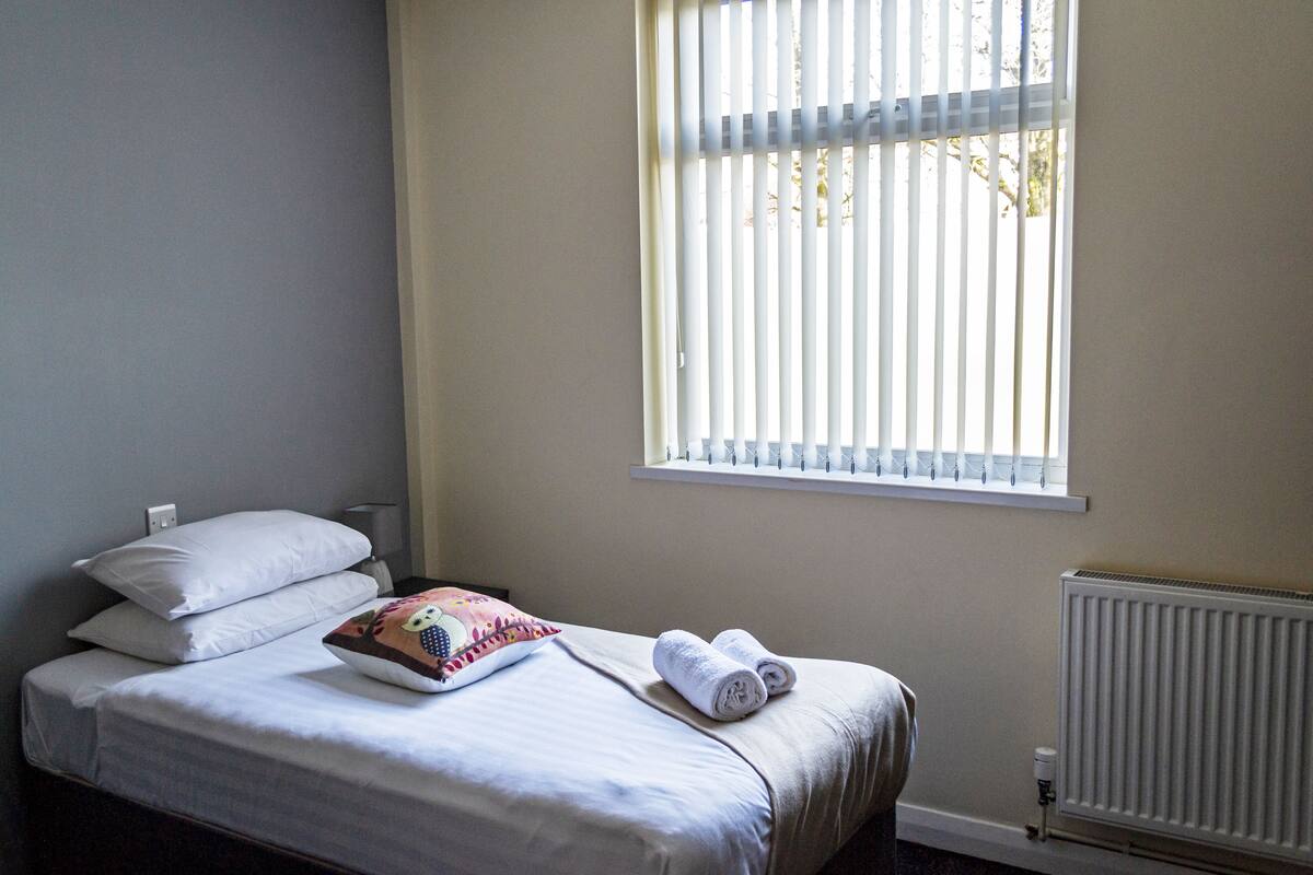 Cama bem arrumada com lençóis brancos, um travesseiro e toalhas dobradas. Há uma janela com persianas abertas parcialmente, permitindo a entrada de luz natural no quarto.