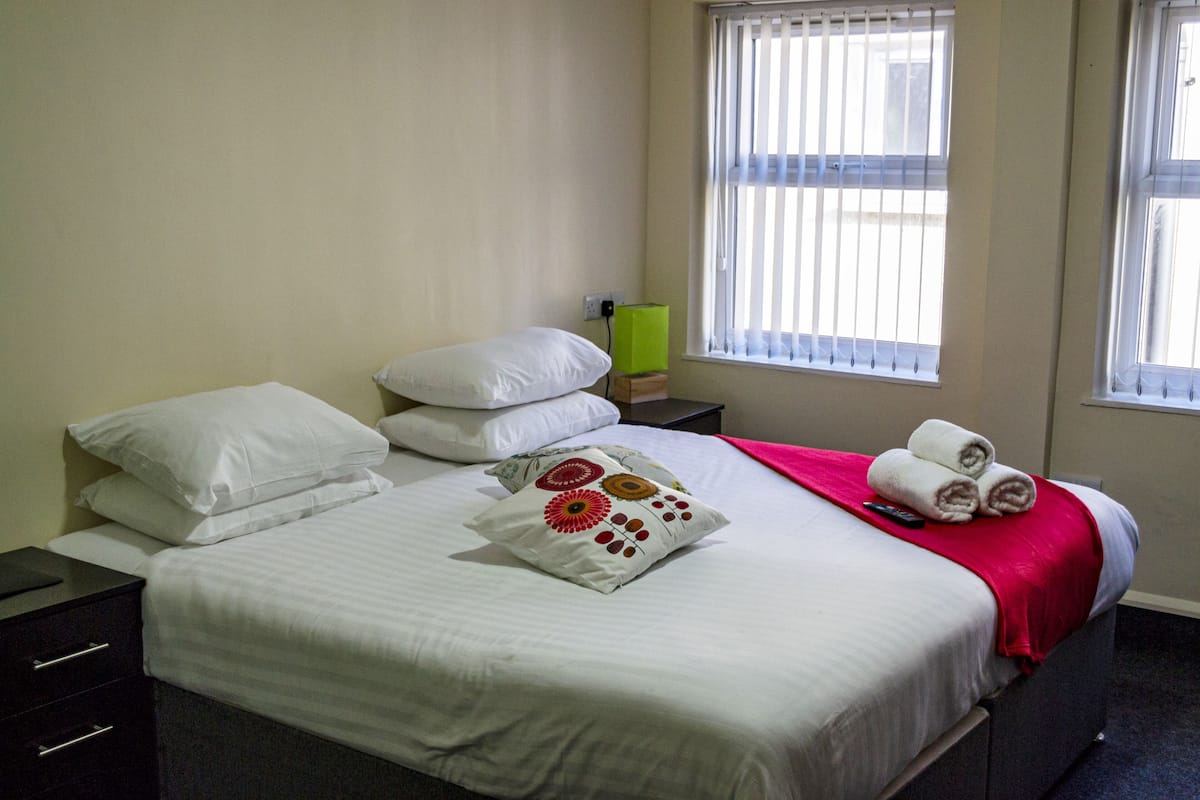 Cama bem arrumada com lençóis brancos, um cobertor vermelho e almofadas decorativas. Há uma mesa de cabeceira com uma luminária e uma caixa verde em cima.