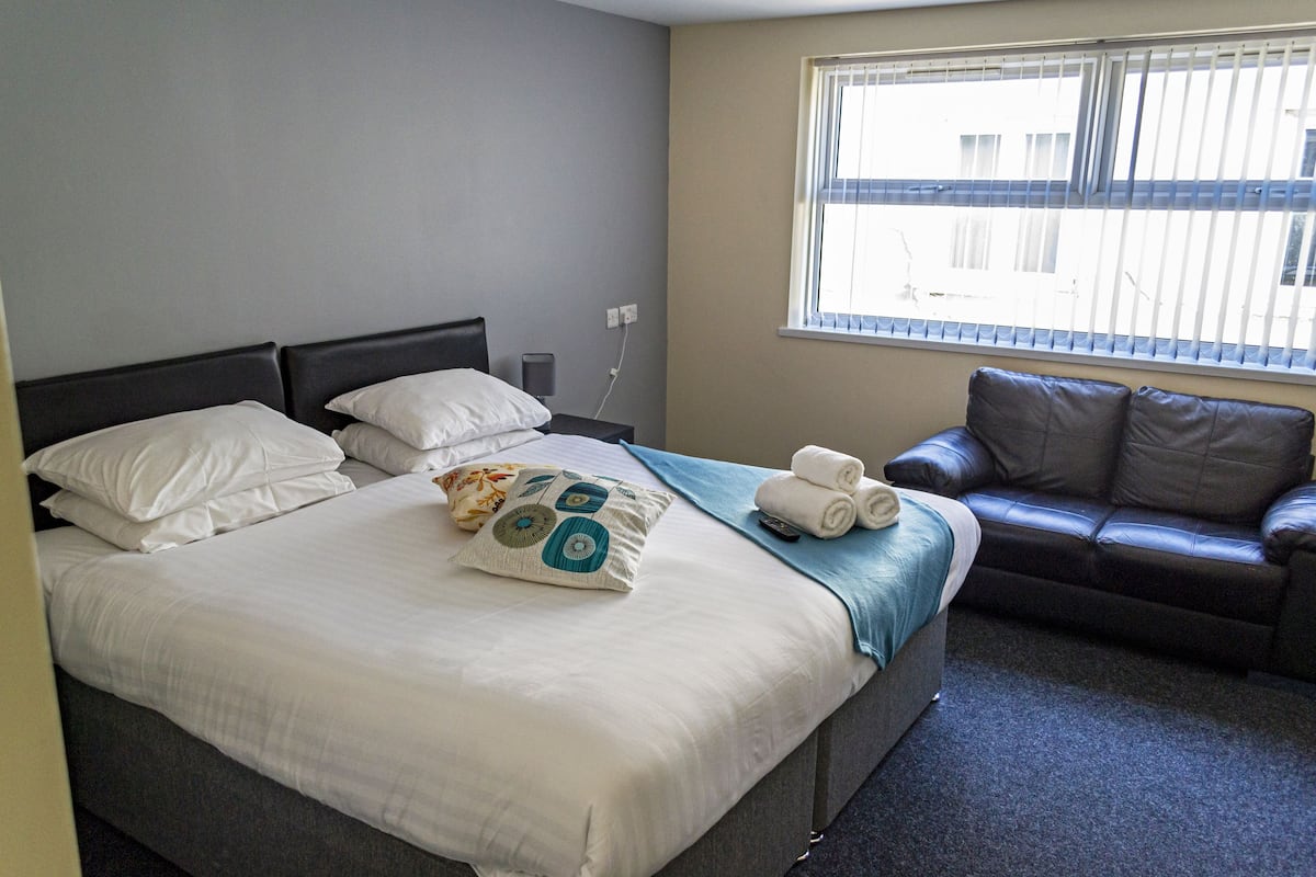 Quarto de hotel com uma cama grande, um sofá de couro marrom e uma janela com persianas.