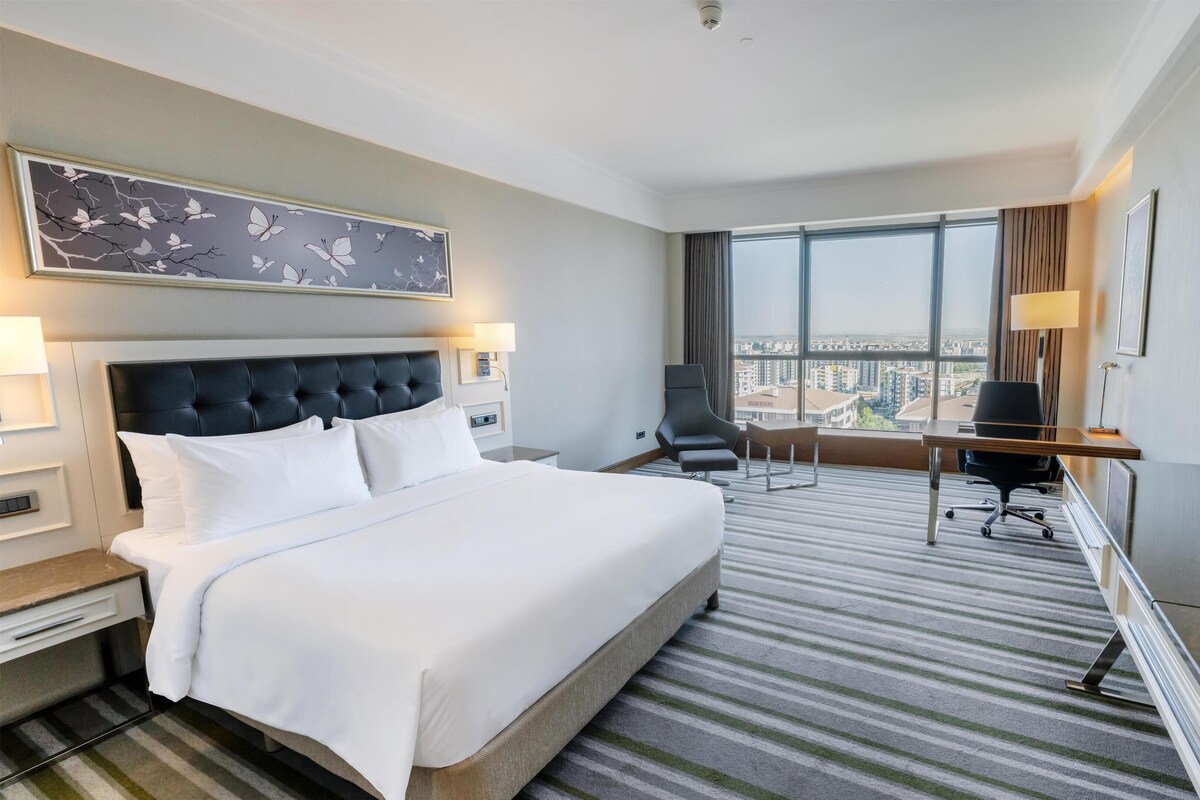 Quarto de hotel com uma cama grande, uma escrivaninha, uma cadeira e vista para a cidade pela janela.