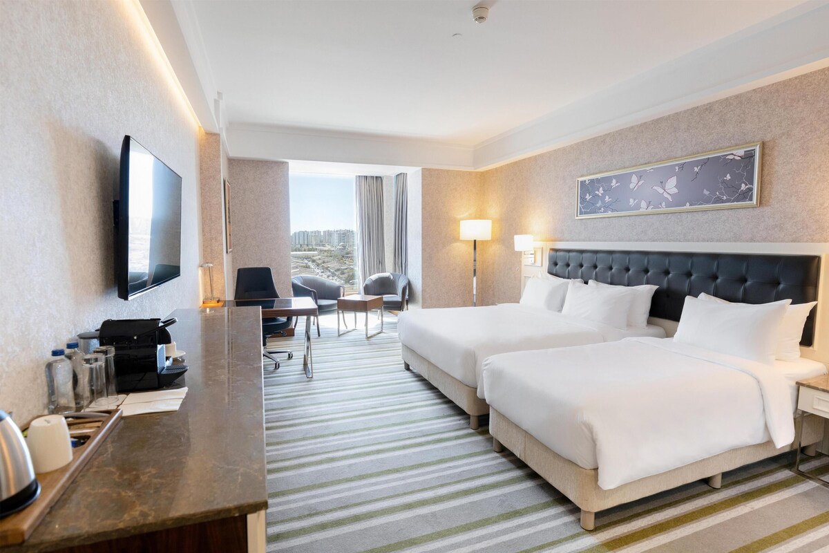 Quarto de hotel com duas camas, uma escrivaninha, uma cadeira e uma televisão.
