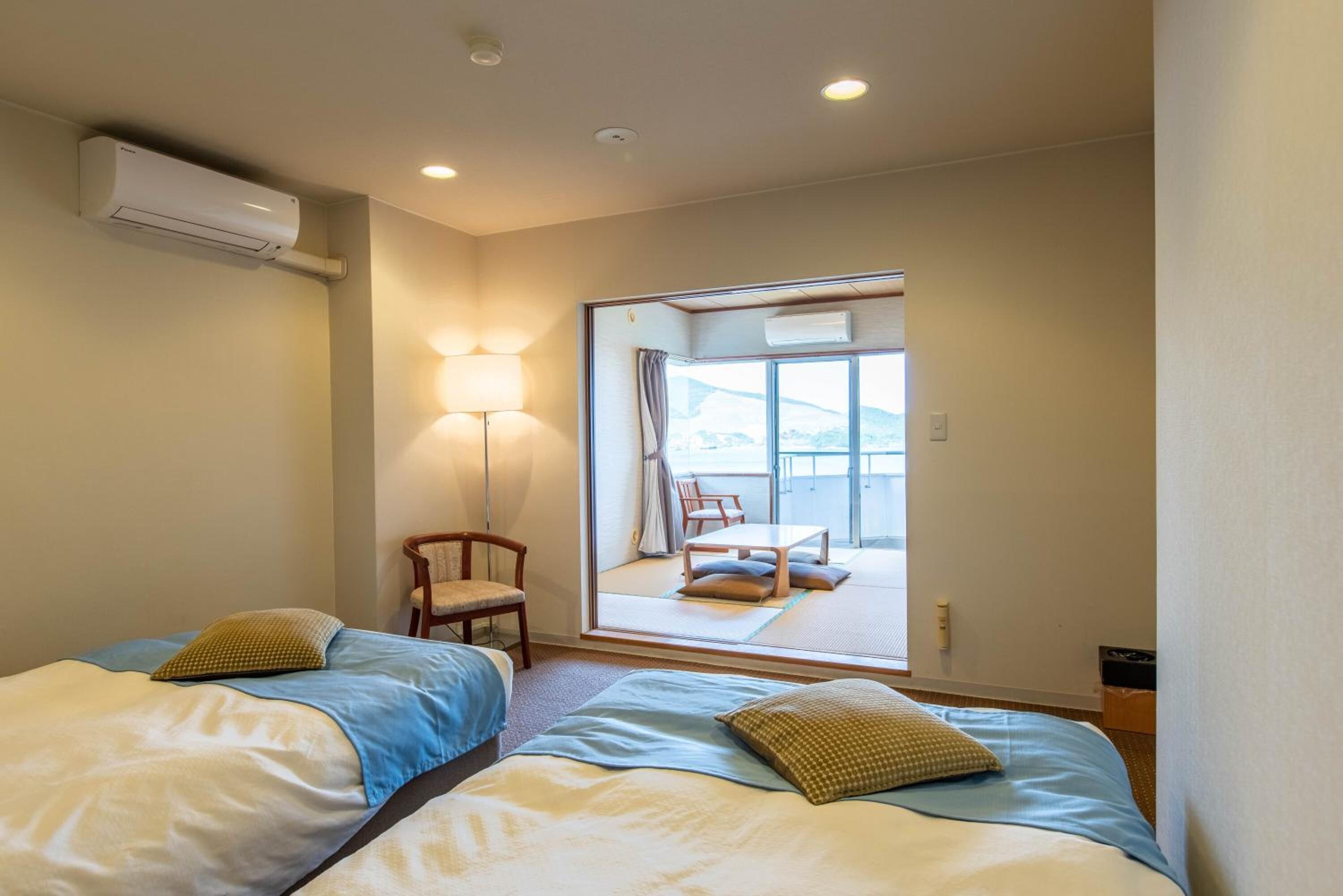 Quarto de hotel com duas camas, uma cadeira, uma luminária e uma porta de vidro deslizante que dá para uma varanda com mesa e cadeiras.