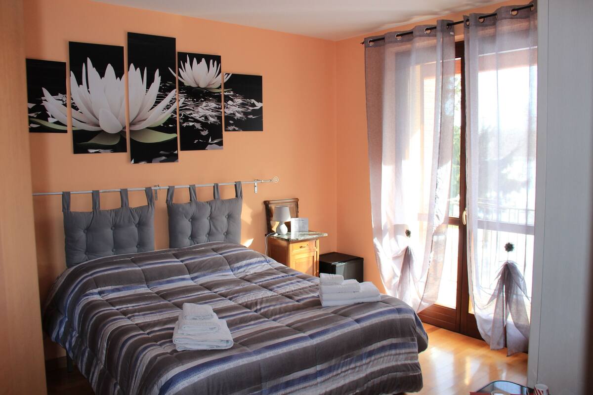 Quarto com uma cama grande, mesa de cabeceira, abajur e janela com cortinas leves.