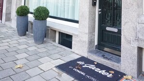 Entrada de hotel com uma grande janela de vidro, uma placa de metal preta sobre a porta e duas plantas em vasos na calçada.