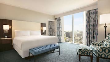 Quarto de hotel com uma cama grande, um banco, uma cadeira e vista para a cidade através de grandes janelas.