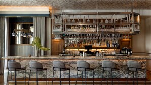 Uma área de bar com bancadas de mármore, diversas prateleiras com garrafas e banquetas.