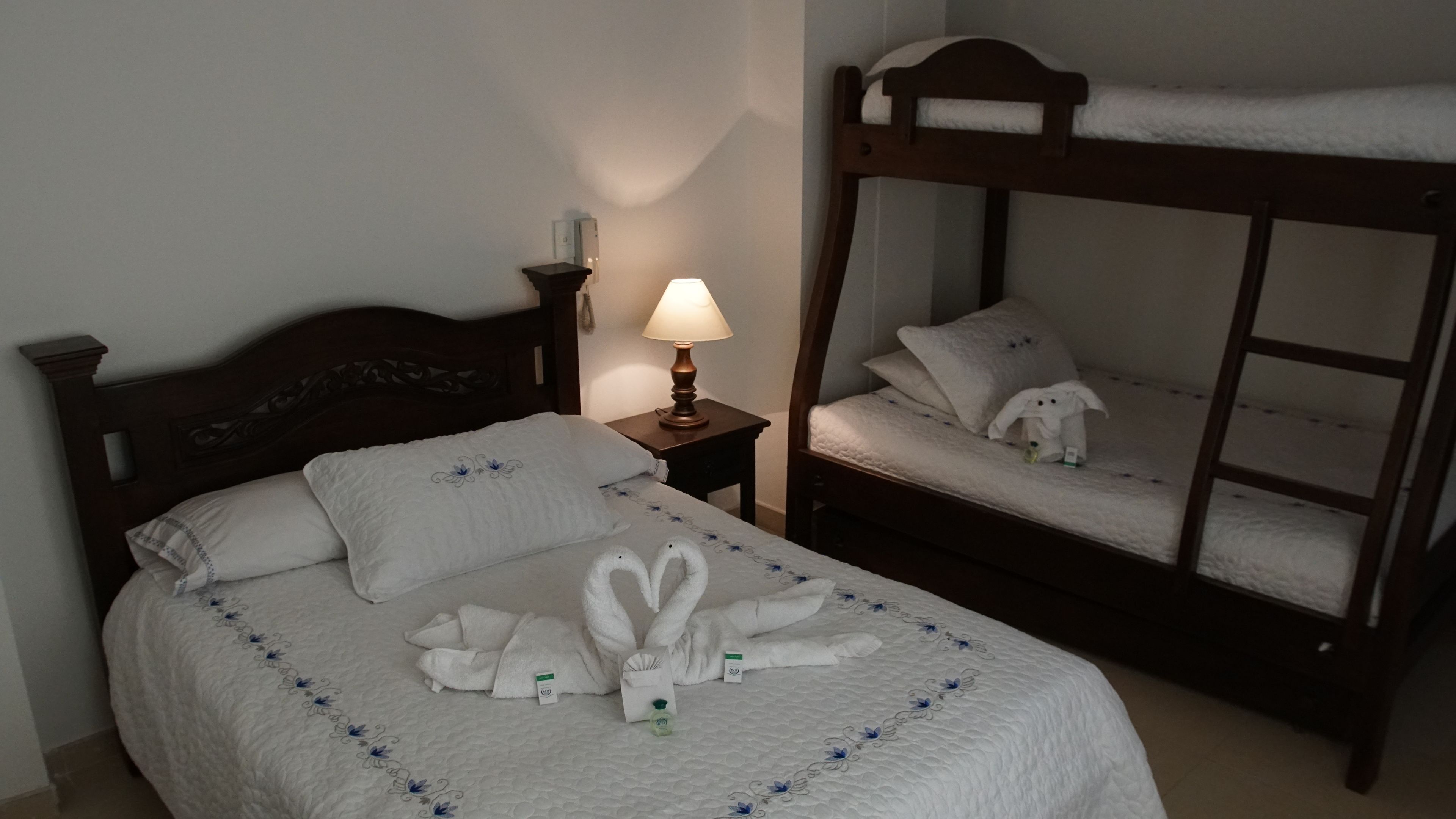 Quarto de hotel com uma cama de solteiro e uma cama beliche, ambos com roupa de cama branca e estampas florais azuis. Há uma mesinha de cabeceira com uma luminária e um pequeno frasco de loção.