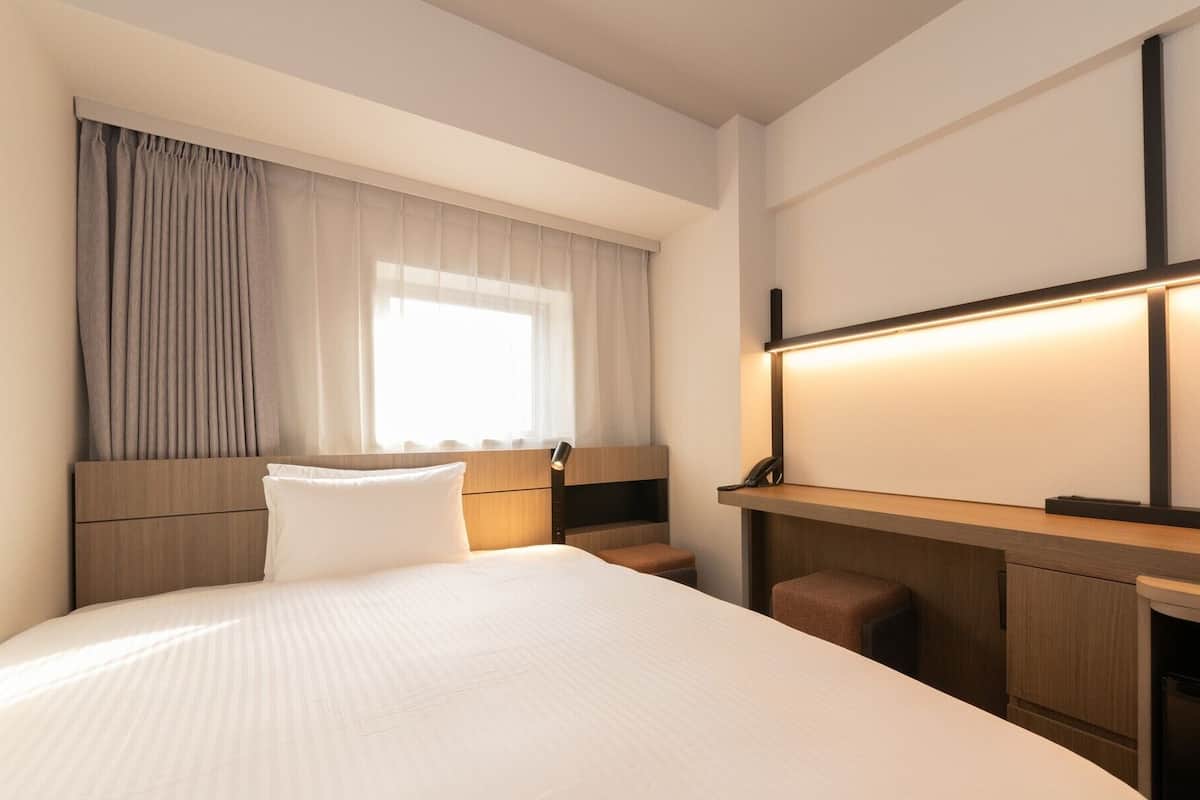 Quarto de hotel com uma cama grande, uma mesinha de cabeceira, uma escrivaninha, uma cadeira, uma janela com cortinas e uma luminária na parede.