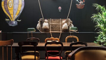 Uma área de refeições com uma mesa alta e cadeiras, uma cesta de vime com sacolas, e um mural na parede com um balão de ar quente.