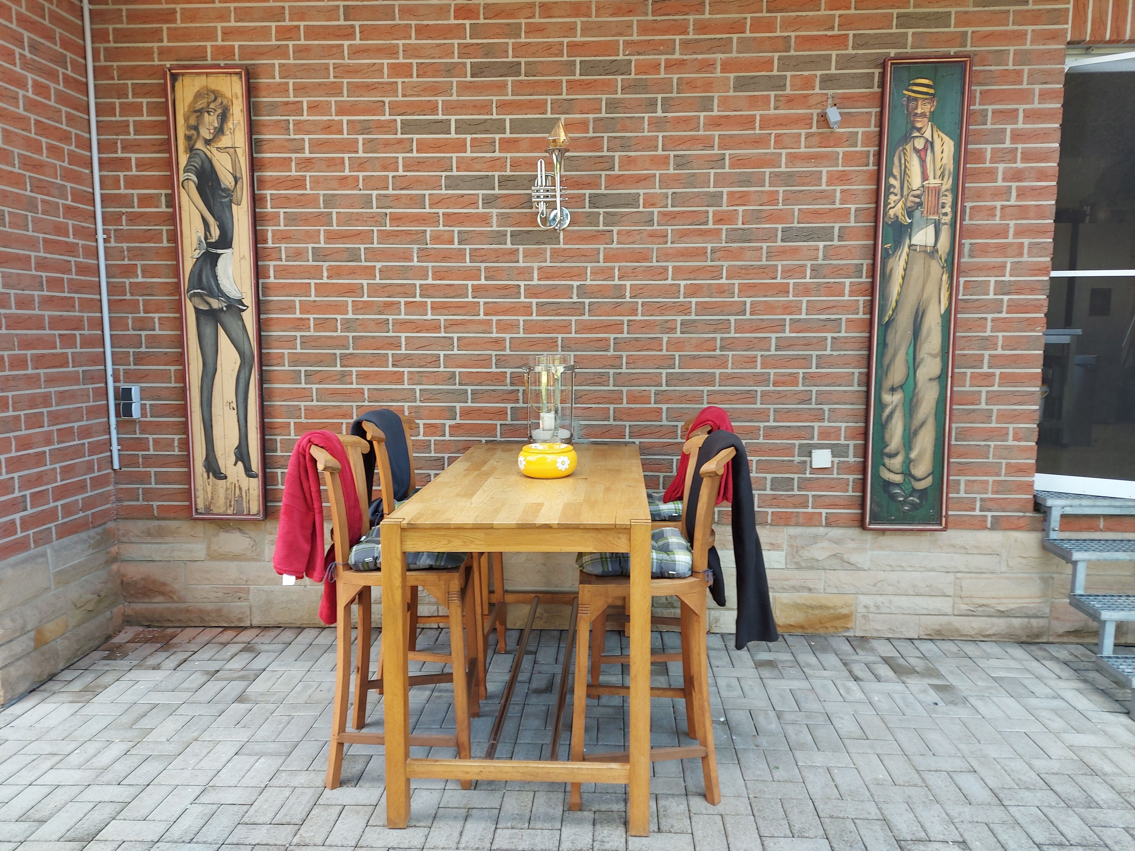 Parede de tijolos com duas pinturas, uma mesa de madeira com cadeiras e uma tigela amarela.