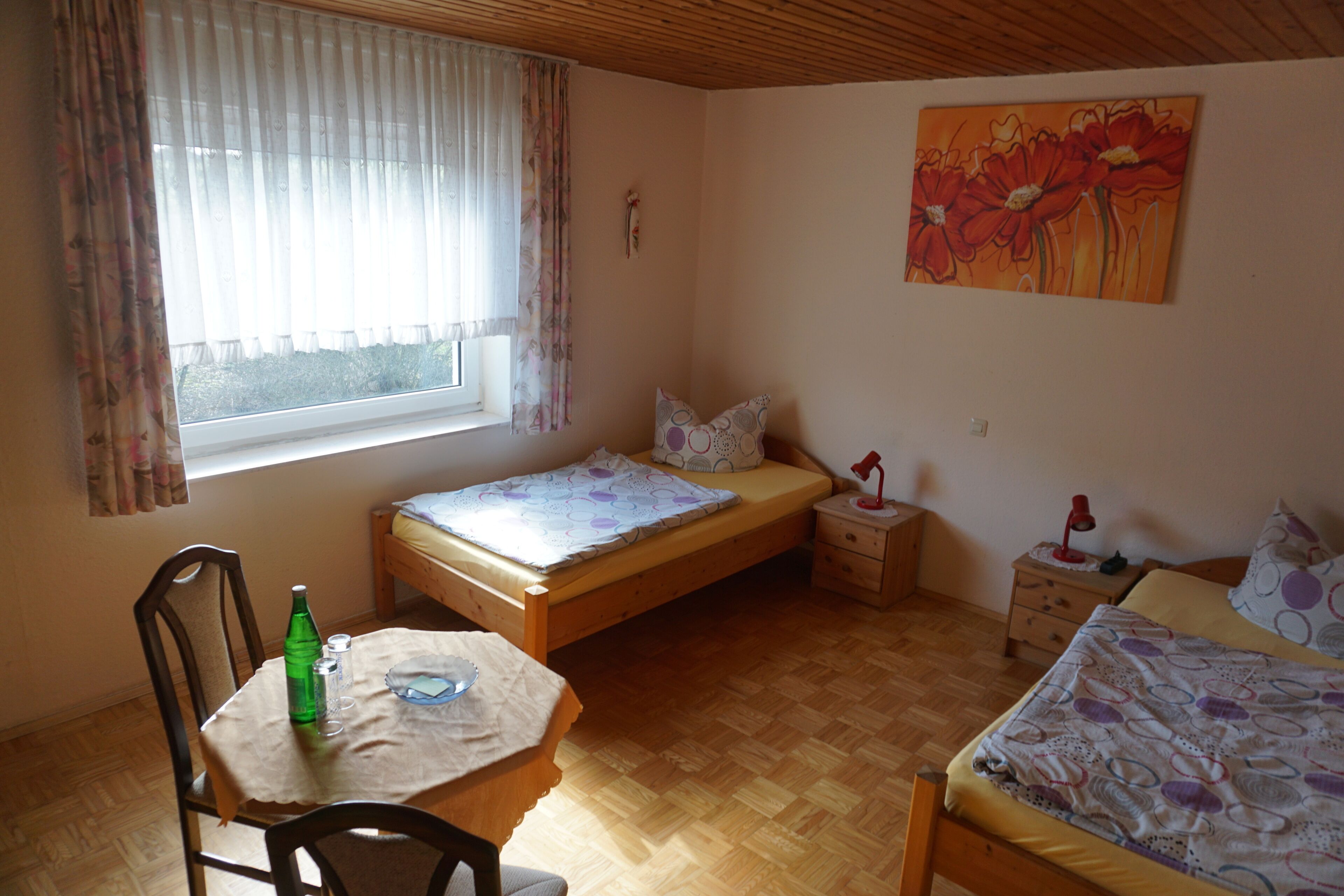 Quarto com duas camas de solteiro, uma mesinha com cadeiras, uma janela com cortinas, um quadro na parede e piso de madeira.