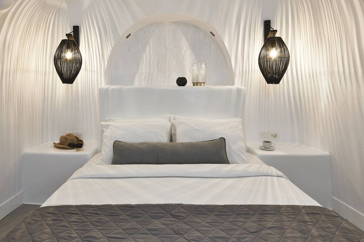 Uma cama bem arrumada com cabeceira acolchoada, duas mesas de cabeceira com uma chávena e um chapéu, e uma luminária na parede.