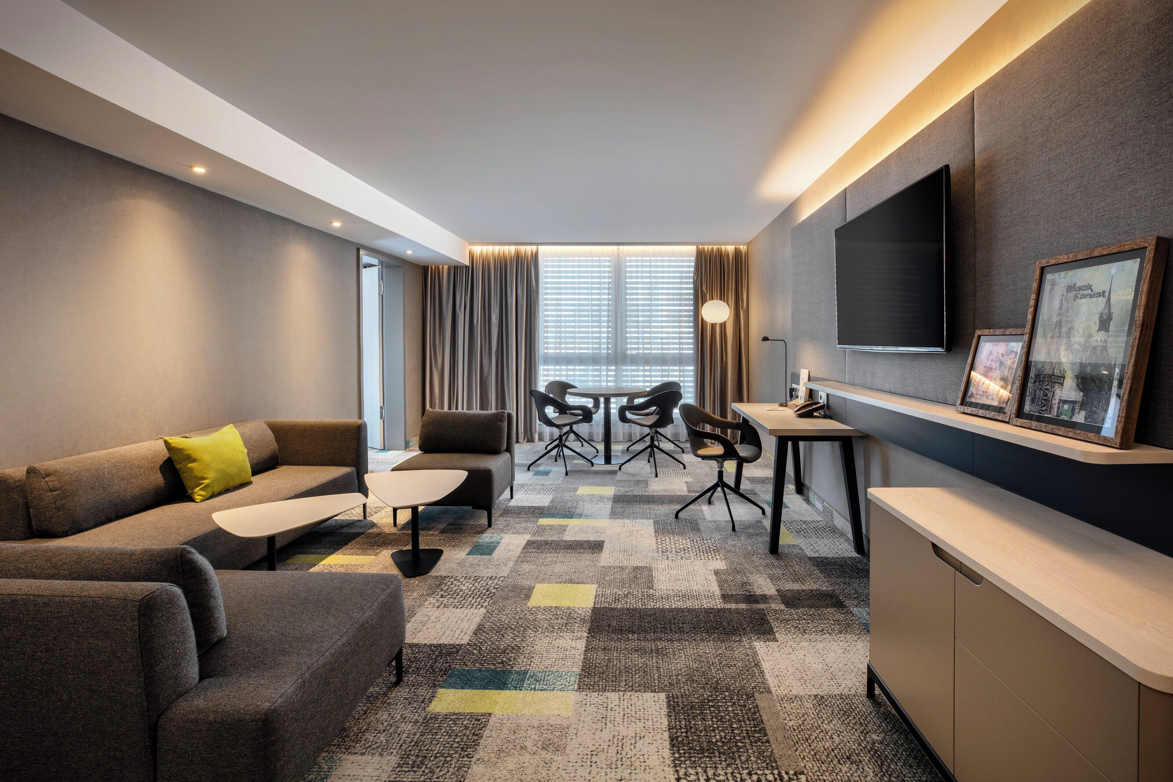 Quarto de hotel moderno com sofá, uma mesinha, escrivaninha, televisão e área de refeições com cadeiras.