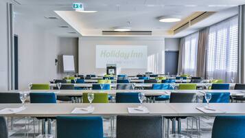 Sala de conferências com mesas e cadeiras, uma tela de projeção exibindo 'Holiday Inn EIN IHG HOTEL' e uma grande janela com cortinas.
