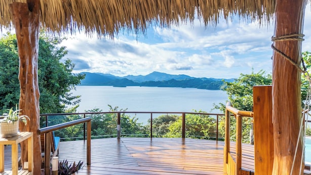 Um deck de madeira com telhado de palha, com vista para um lago e montanhas.