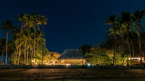 Uma cabana à beira-mar com telhado de palha, rodeada por palmeiras e uma guirlanda de luzes.