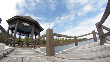 Um píer de madeira que leva a um gazebo com uma placa, cercado por água e palmeiras.