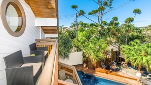 Uma área à beira da piscina com móveis de vime, um deck de madeira e vista para um jardim exuberante com árvores e um céu azul límpido.
