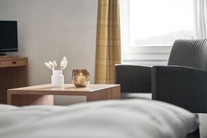 Quarto de hotel com cama, mesa de cabeceira com um vaso e vela, televisão e janela com cortinas.