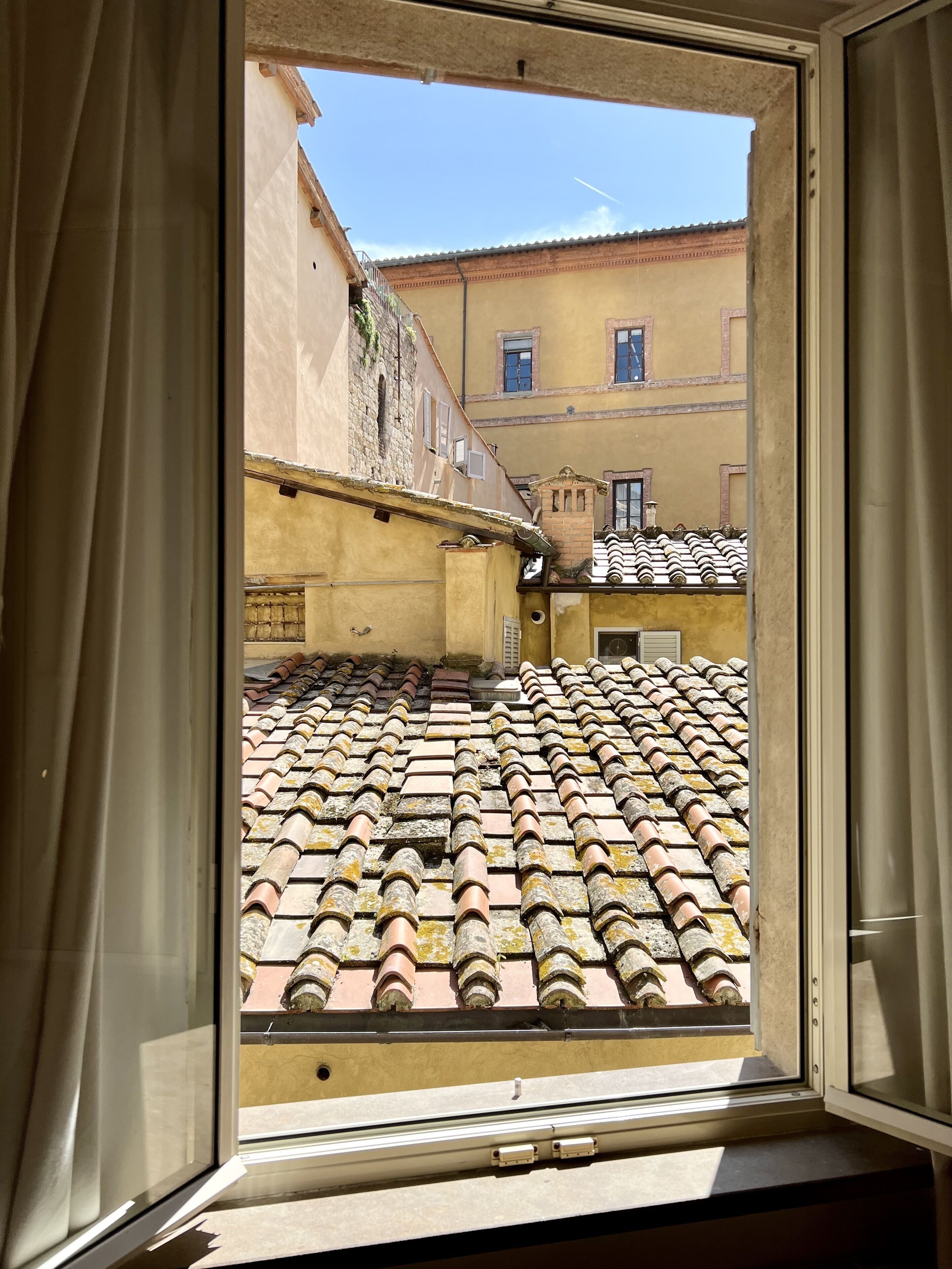 Vista pela janela de um edifício com telhados de terracota e fachada amarela.