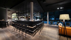 Uma área de bar moderna com uma ilha central, banquetas e um espaço de estar com um sofá e cadeiras.
