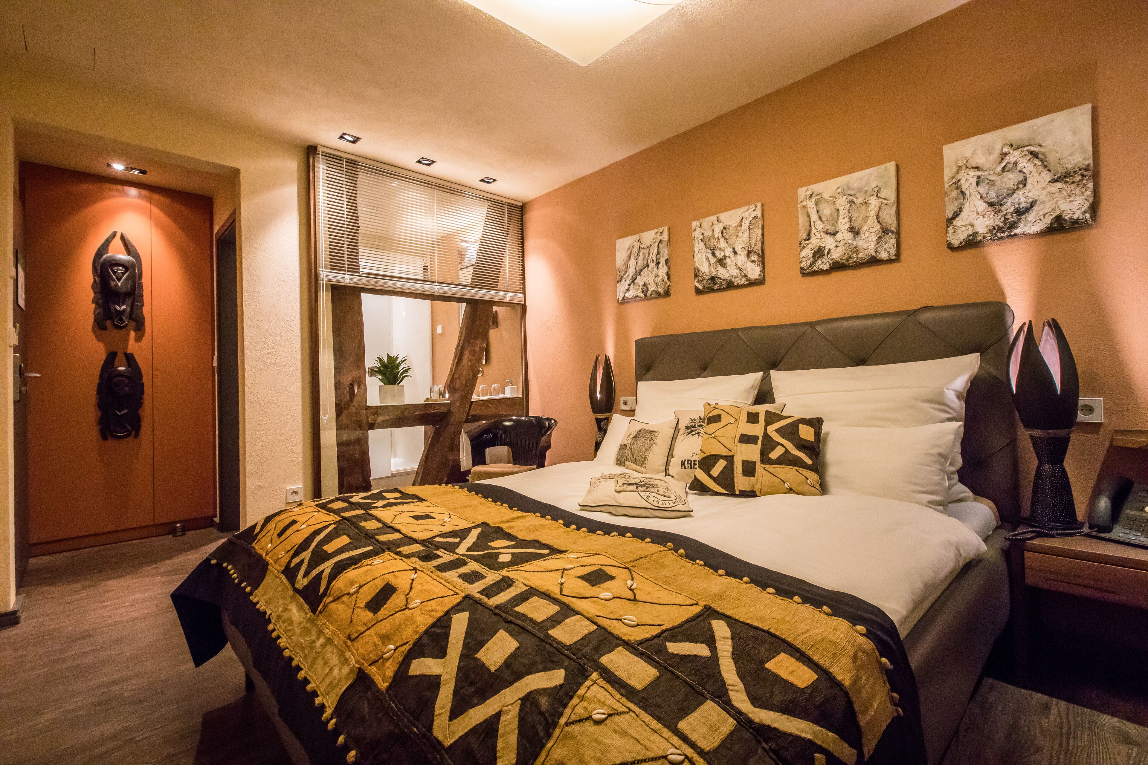 Quarto com uma cama grande, mesa de cabeceira, escrivaninha e cadeira. Há obras de arte nas paredes acima da cama e uma porta com um objeto decorativo pendurado.