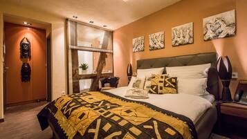 Quarto com uma cama grande, mesa de cabeceira, escrivaninha e cadeira. Há obras de arte nas paredes acima da cama e uma porta com um objeto decorativo pendurado.